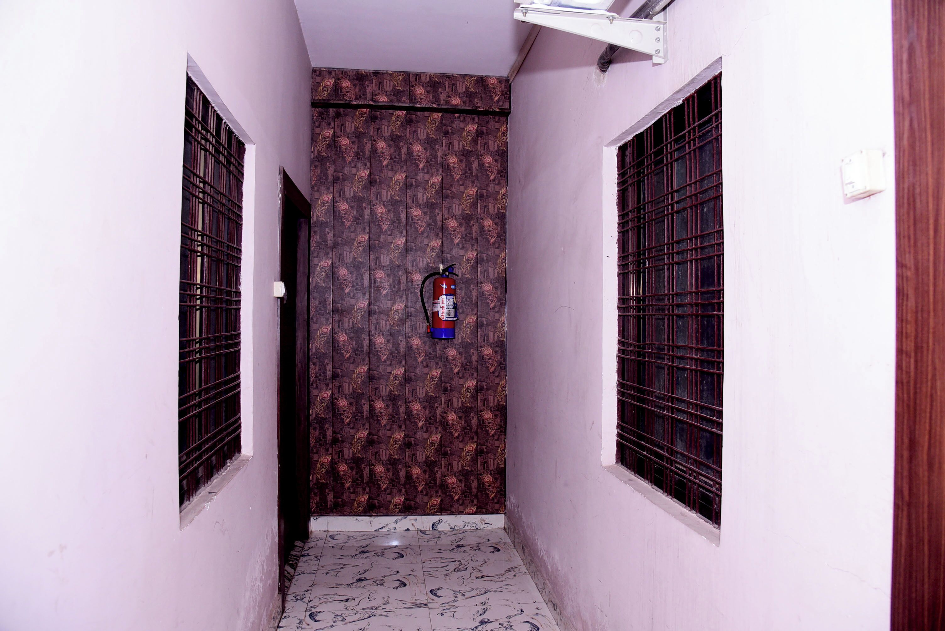 Um corredor estreito com papel de parede estampado, janelas com grades e um extintor fixado na parede.