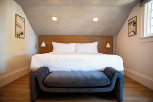 Quarto com uma cama grande, cabeceira de madeira, duas luminárias de apoio e um pequeno banco.