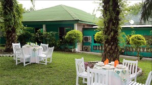 Uma área de refeições ao ar livre com cadeiras e mesas brancas dispostas em um gramado, rodeada por vegetação e um edifício com telhado verde.