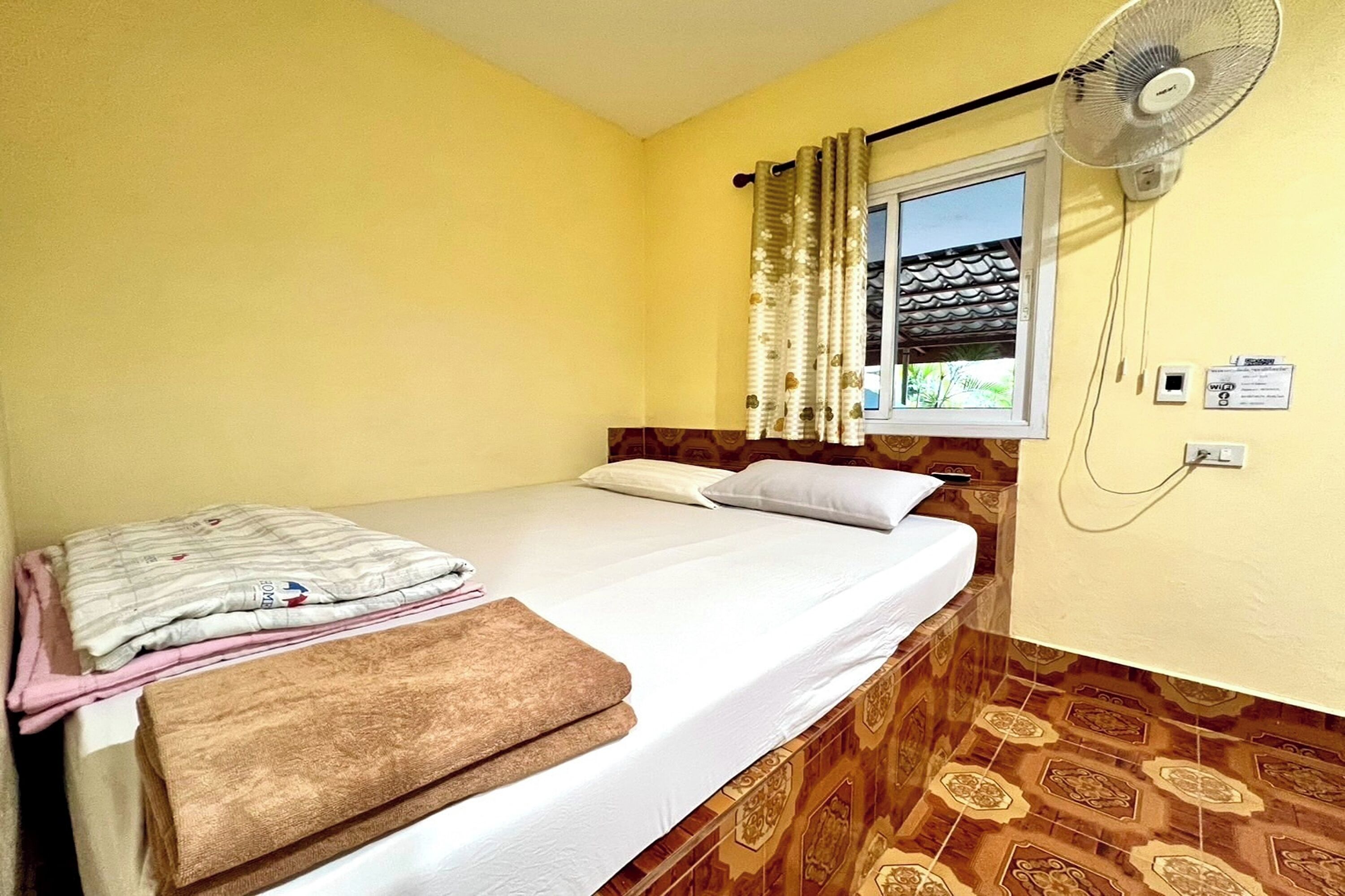Uma cama de solteiro com colchão branco, duas almofadas e uma toalha castanha. Uma janela com cortinas estampadas é visível, e há um ventilador fixado na parede.