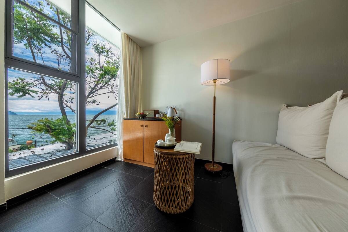 Um quarto com uma janela grande, um sofá, um abajur e uma pequena mesa com um vaso.