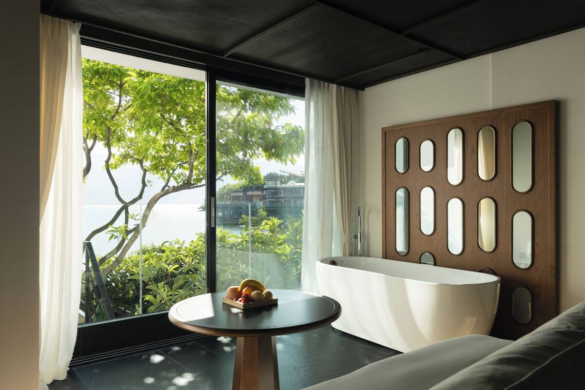 Uma casa de banho moderna com uma banheira independente, uma mesa redonda de madeira com fruta, e vista para um corpo de água através de grandes janelas.