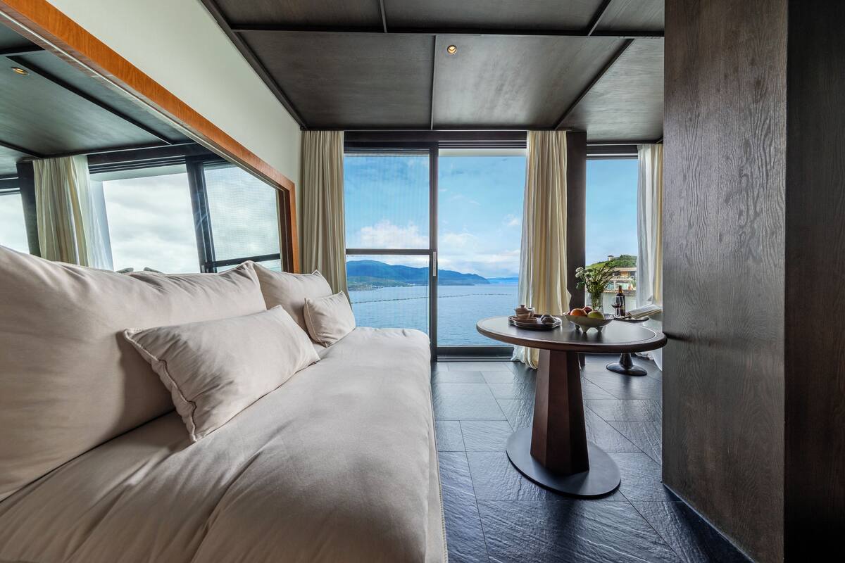 Quarto de hotel moderno com uma cama grande, uma mesa redonda com um vaso e frutas, e vista para o mar e montanhas através de amplas janelas.