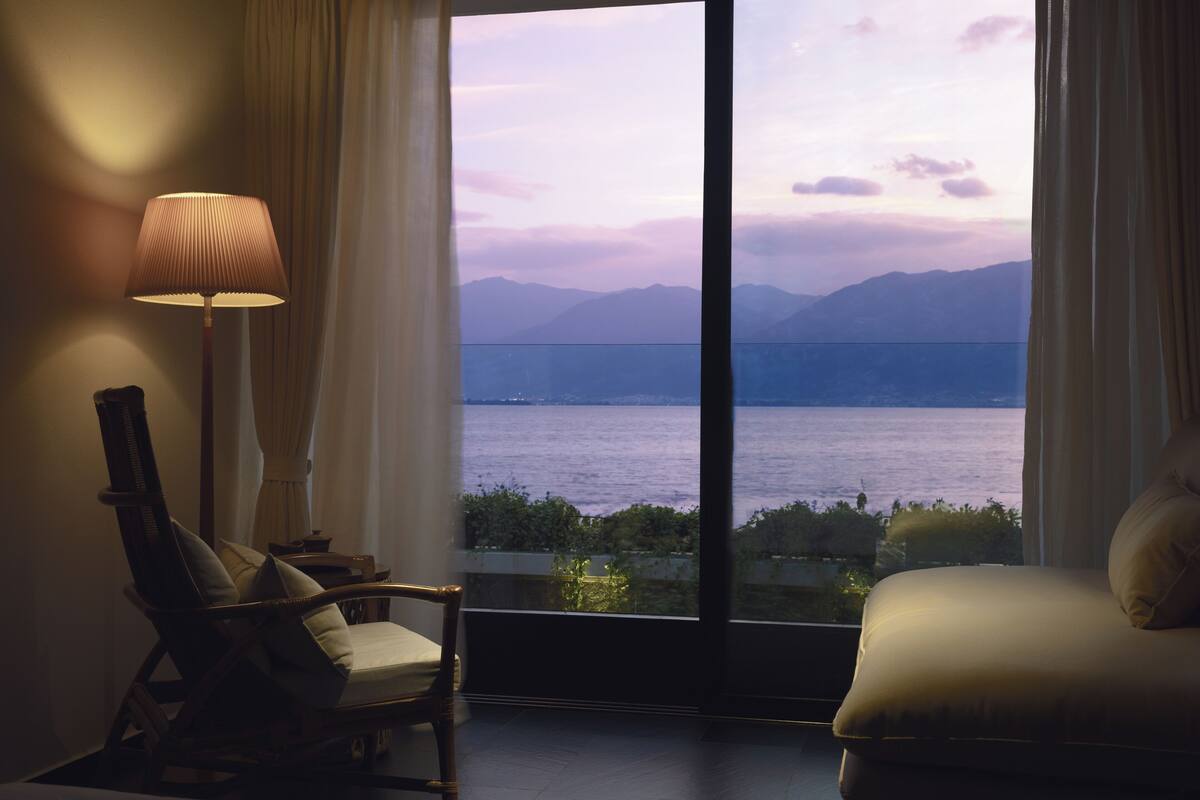 Quarto com vista para um lago e montanhas, uma cadeira, uma cama e um abajur.