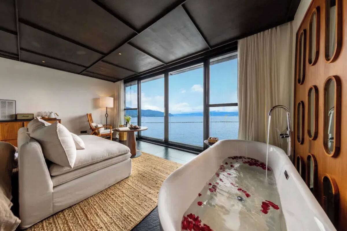 Quarto de hotel moderno com uma janela grande, uma banheira com pétalas de rosa, um sofá e um painel de parede de madeira.