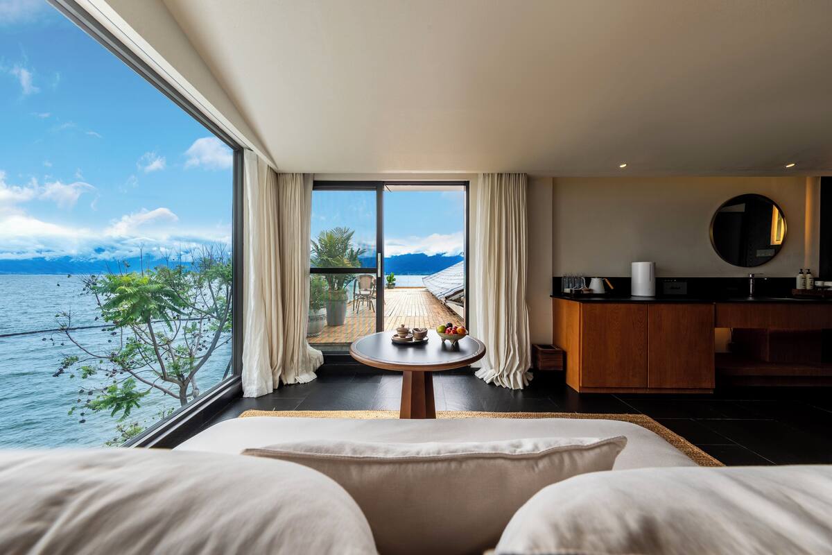 Quarto de hotel com uma grande janela, uma cama, uma mesa pequena com uma tigela de frutas e vista para o mar e montanhas.