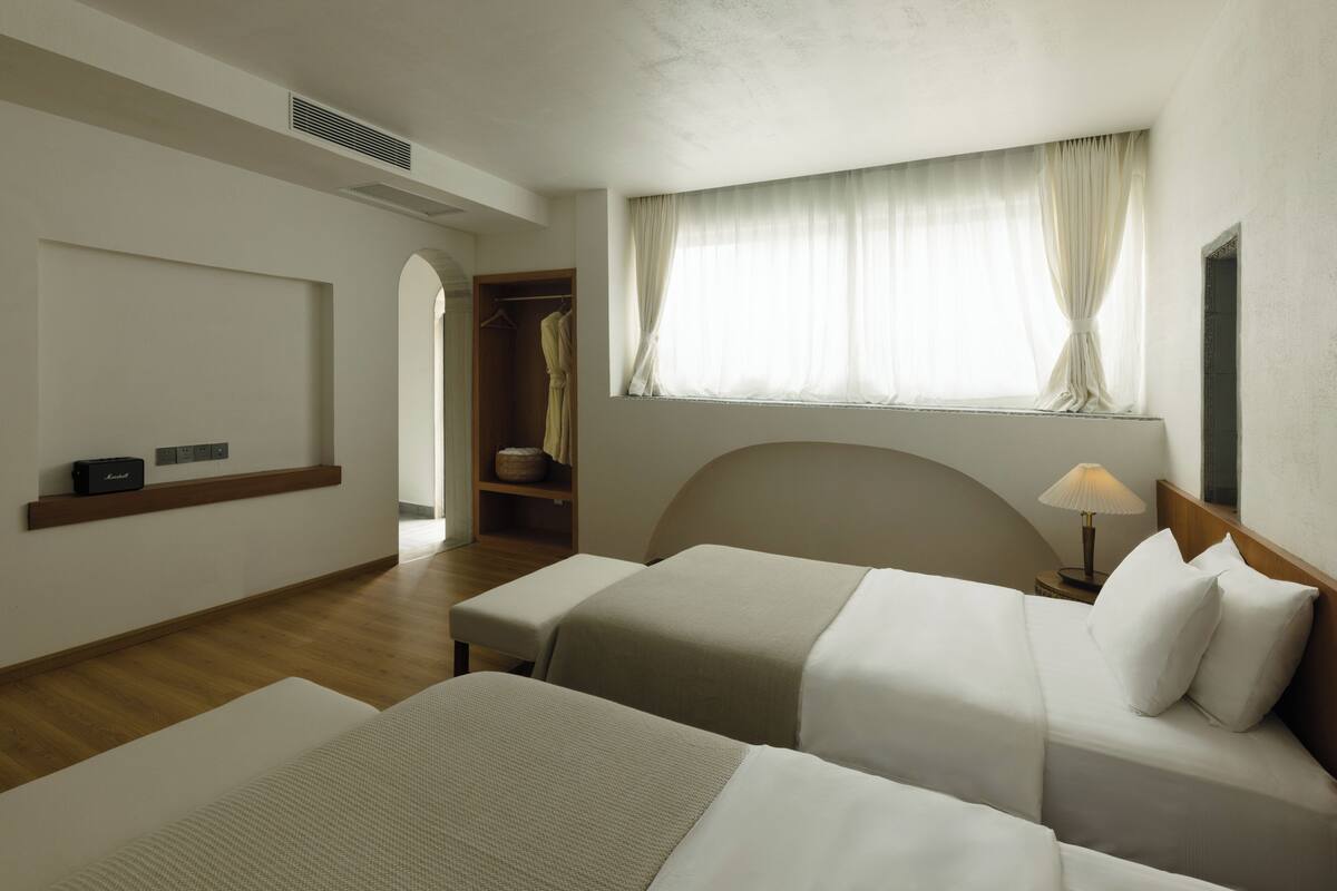 Quarto de hotel com duas camas, uma mesa de cabeceira com abajur, um guarda-roupa e uma janela com cortinas.