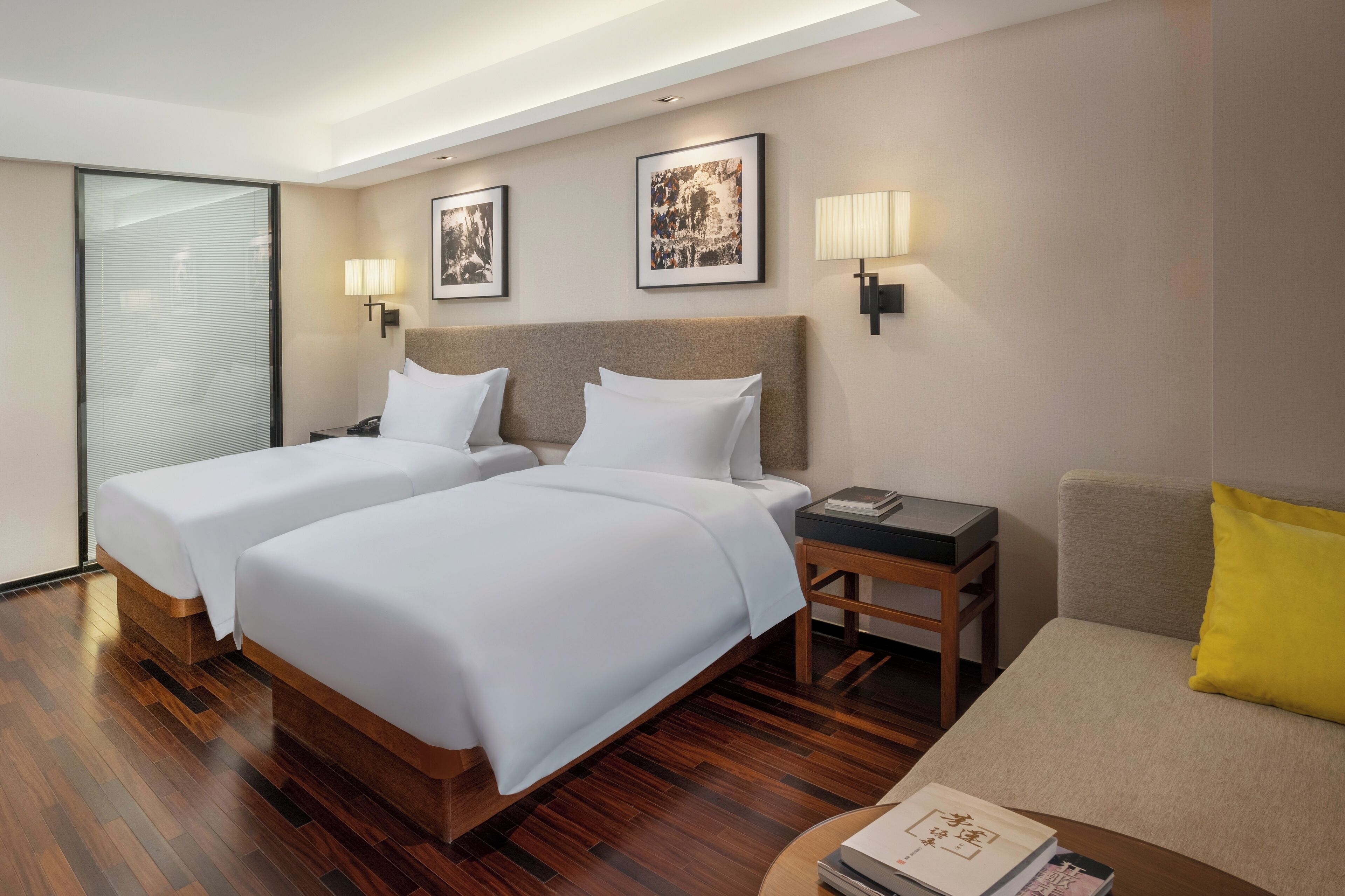 Quarto de hotel com duas camas, um sofá, uma mesinha e duas luminárias fixadas na parede.