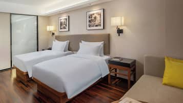 Quarto de hotel com duas camas, um sofá, uma mesinha e duas luminárias fixadas na parede.