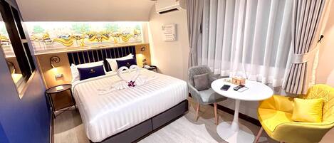 Quarto de hotel com uma cama, uma mesinha redonda, uma cadeira amarela, um poltrona cinza e vista para um jardim pela janela.