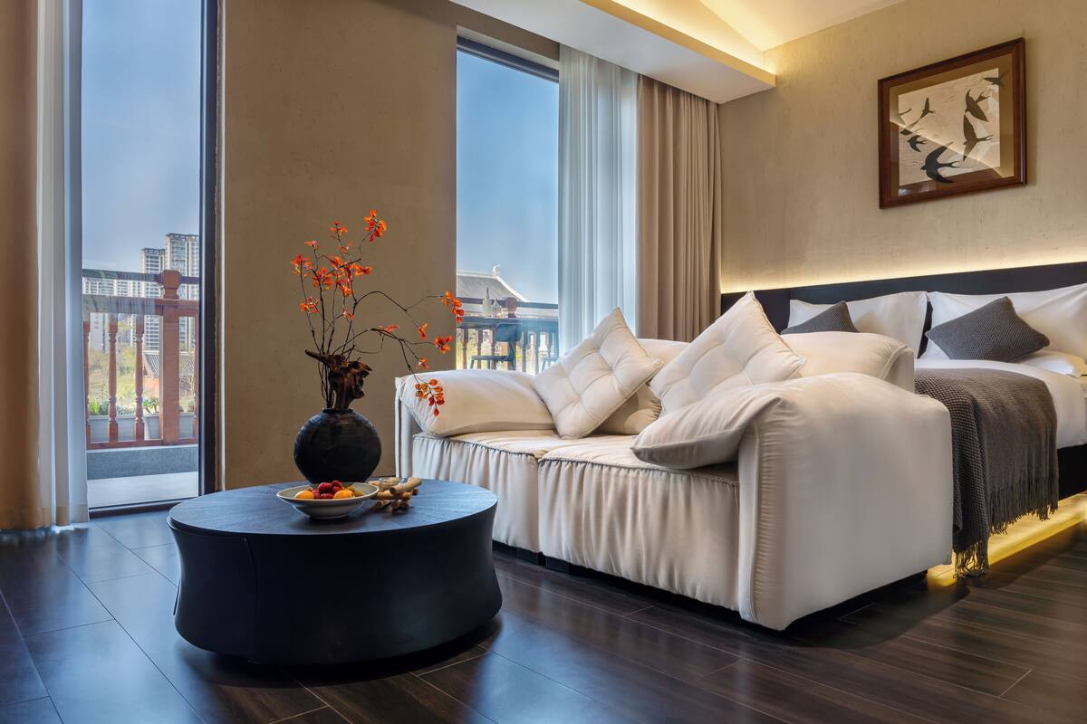 Quarto de hotel moderno com uma janela grande, uma mesa redonda preta, um sofá-cama e uma obra de arte emoldurada na parede.