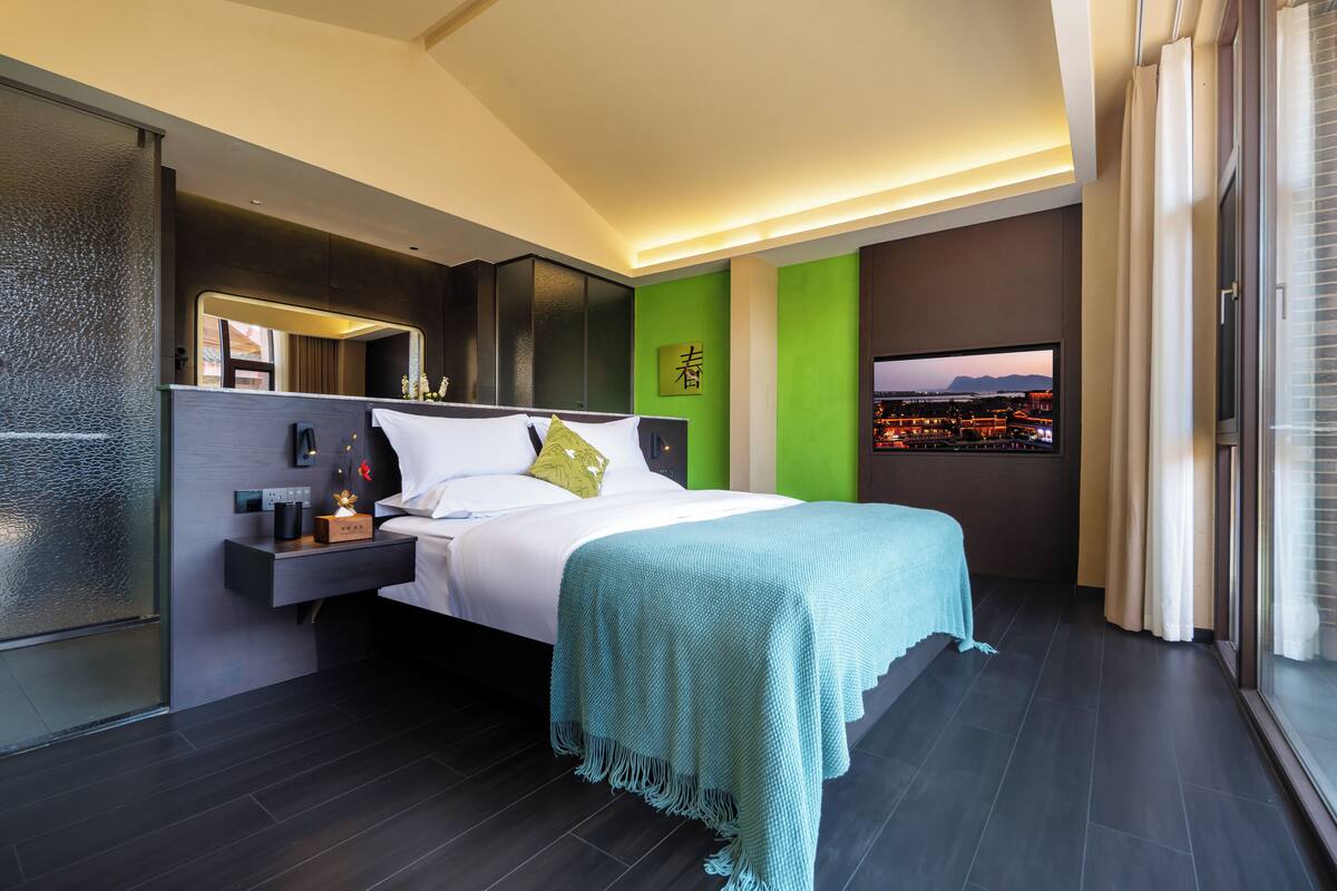 Quarto de hotel moderno com uma cama grande, uma mesa de cabeceira, um espelho, uma parede de destaque verde e vista para a cidade.