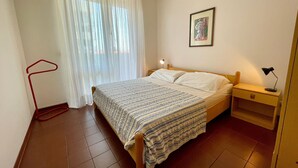 Quarto com cama, mesa de cabeceira, abajur, quadro na parede e janela com cortinas.