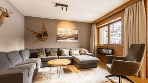 Uma sala de estar moderna com um sofá modular cinza, uma mesa de centro redonda e uma poltrona cinza. Uma janela grande com cortinas permite a entrada de luz natural, e um abajur fixado na parede oferece iluminação adicional.