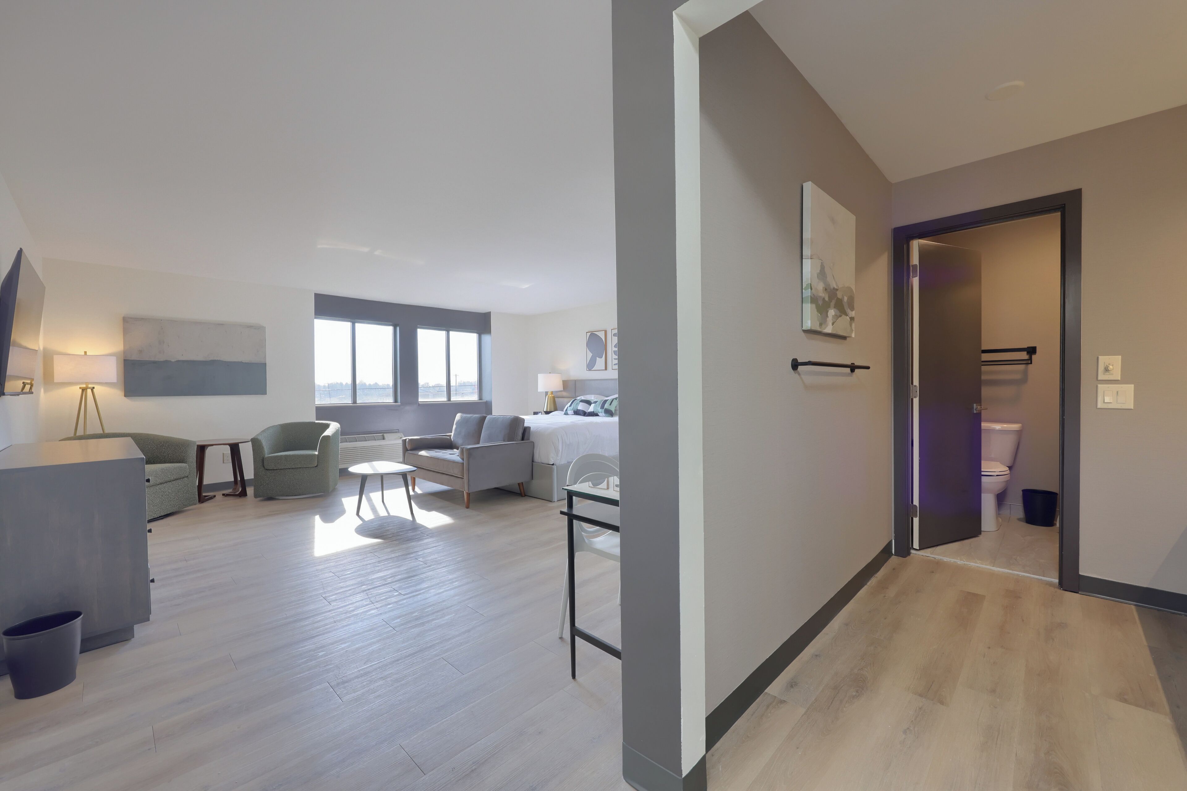 Quarto de hotel moderno com uma cama grande, área de estar com sofá e cadeiras, uma mesa pequena e vista da cidade através de janelas amplas.