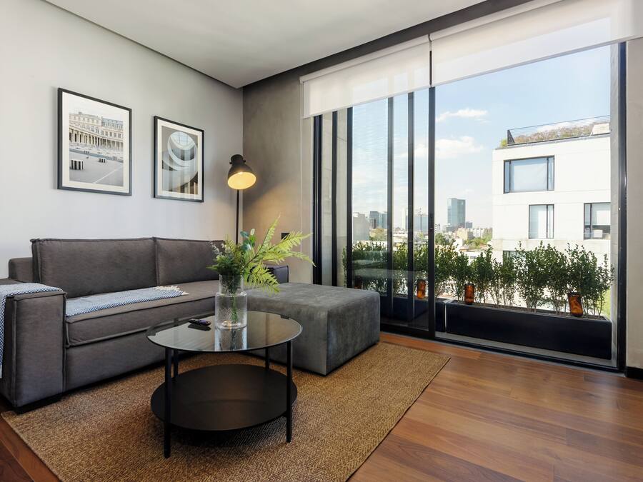 Uma sala de estar moderna com um sofá cinza, uma mesa de centro de vidro e uma janela grande com vista para a cidade.