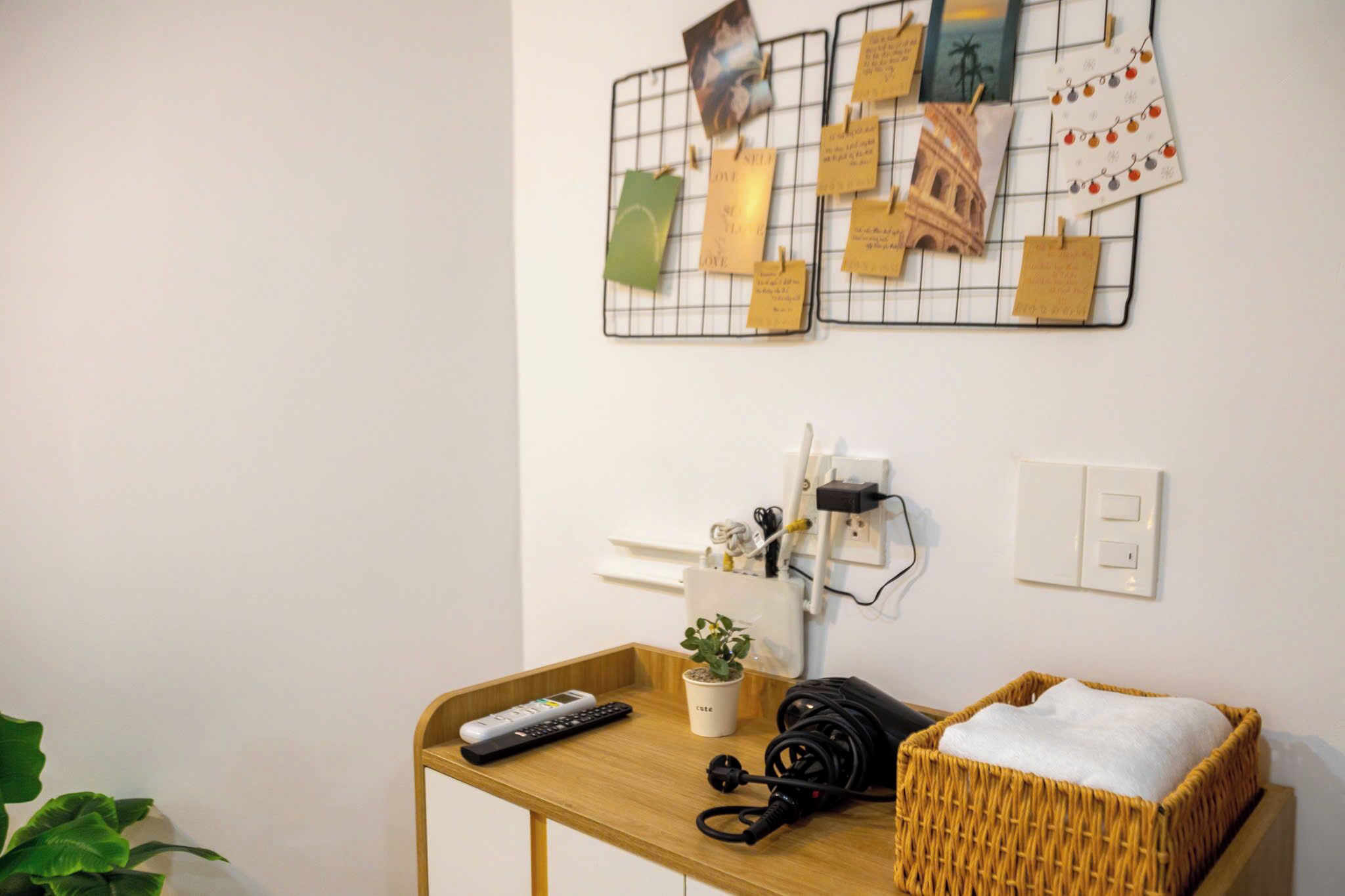 Quarto com mesa de madeira contendo um controle remoto, uma planta e uma cesta com toalha. Uma estação de carregamento na parede e uma tomada elétrica são visíveis.