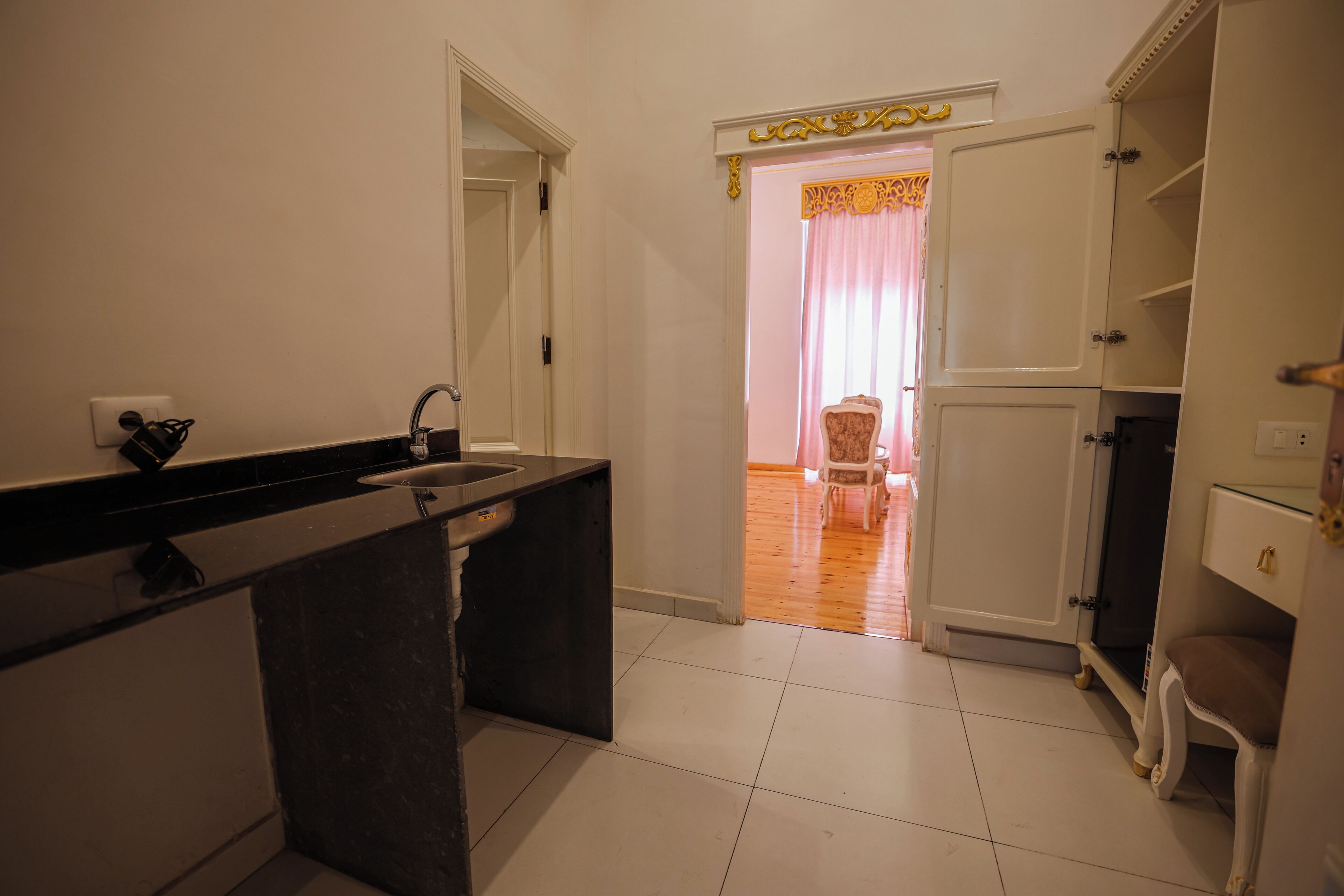 Casa de banho com lavatório, espelho e armários. Há uma porta que dá para uma divisão com uma cadeira e uma mesa.