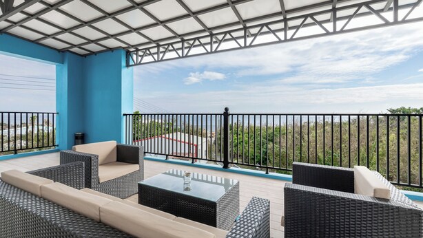 Uma área de estar coberta ao ar livre, com móveis de vime e uma mesa de vidro, com vista para um jardim paisagístico.