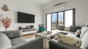 Uma sala de estar moderna com televisão de tela plana, sofás cinzas, uma mesa de centro e uma janela grande com vista para edifícios.
