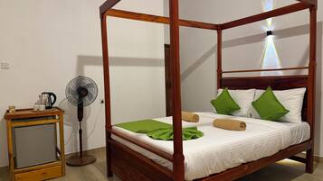 Uma cama de dossel com estrutura de madeira, roupa de cama branca e almofadas verdes. Um ventilador está posicionado ao lado da cama, e uma pequena mesa com chaleira e xícaras é visível.