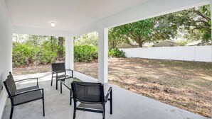 Um alpendre coberto com duas cadeiras pretas e uma mesa, cercado por um muro branco e vegetação.