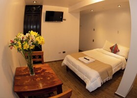 Quarto de hotel com cama, televisão na parede, mesa de madeira com um vaso de flores e uma pequena escrivaninha.