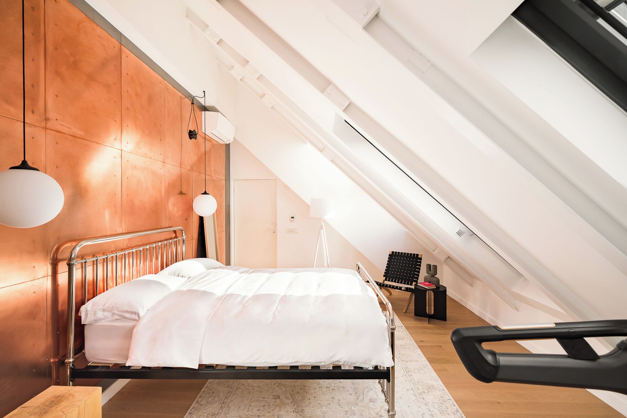 Quarto moderno com teto inclinado, estrutura de cama de metal e esteira no canto.