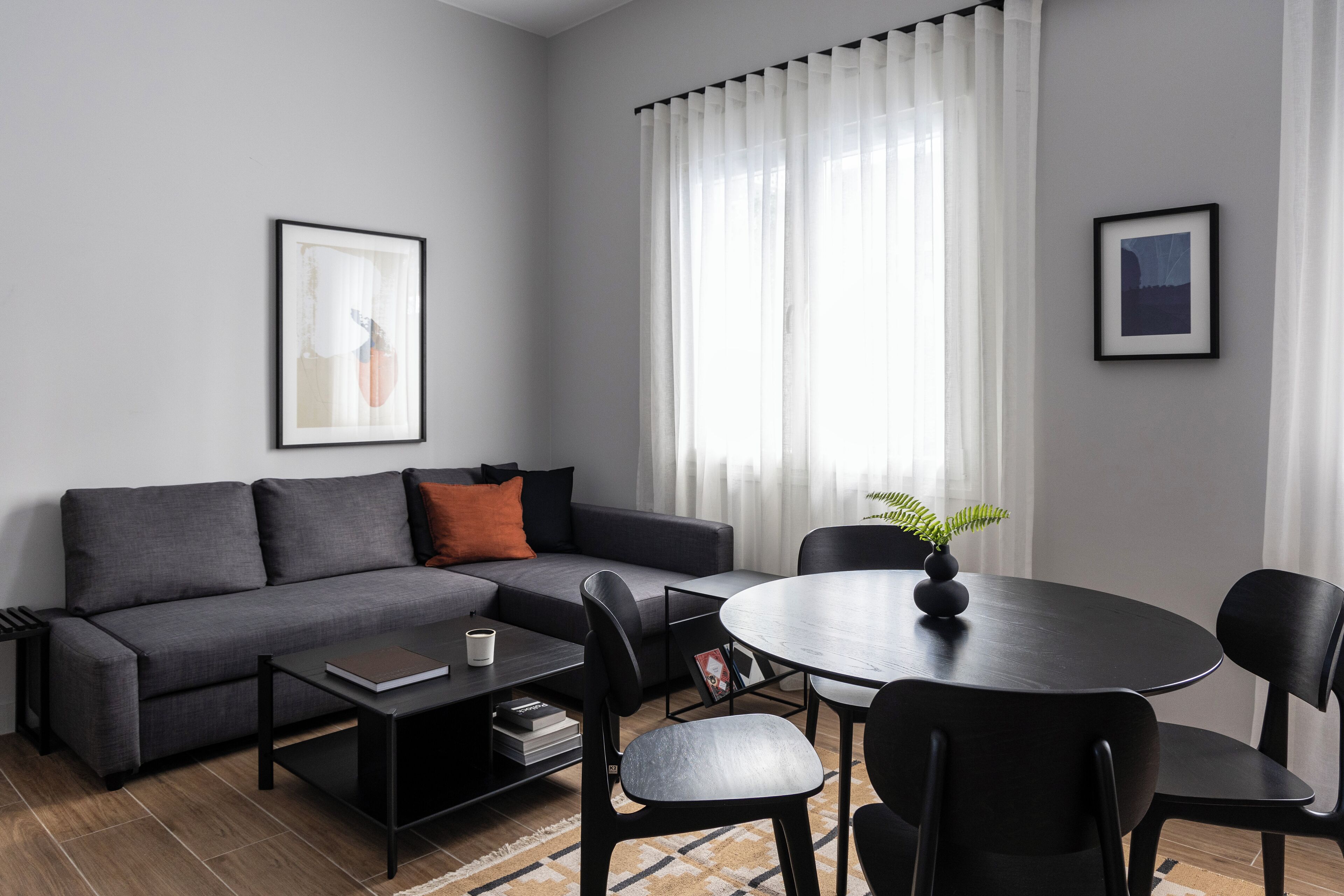 Uma sala de estar moderna com um sofá cinza, mesa de centro preta e uma mesa de jantar redonda com cadeiras pretas. Há um quadro emoldurado na parede e um vaso com planta na mesa.