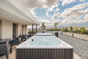 Um terraço no telhado com duas banheiras de hidromassagem, uma área de refeições com cadeiras e mesa, e vista para um horizonte urbano com montanhas ao fundo.