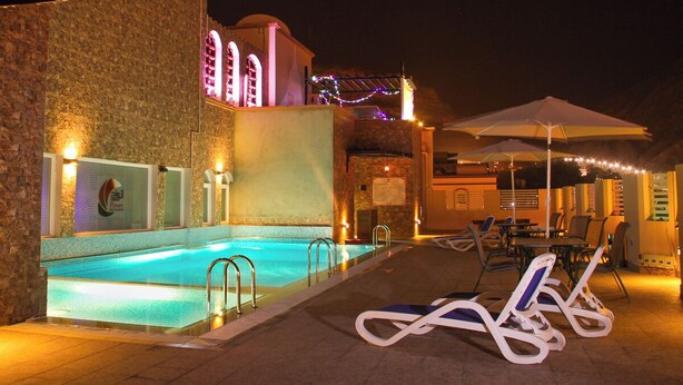 Uma área de piscina ao ar livre com espreguiçadeiras e guarda-sóis, cercada por paredes de pedra e um edifício com janelas iluminadas.