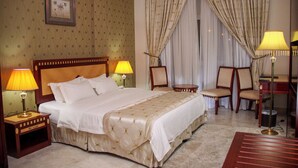 Quarto de hotel com uma cama grande, duas luminárias laterais, uma cadeira, uma escrivaninha e um abajur.