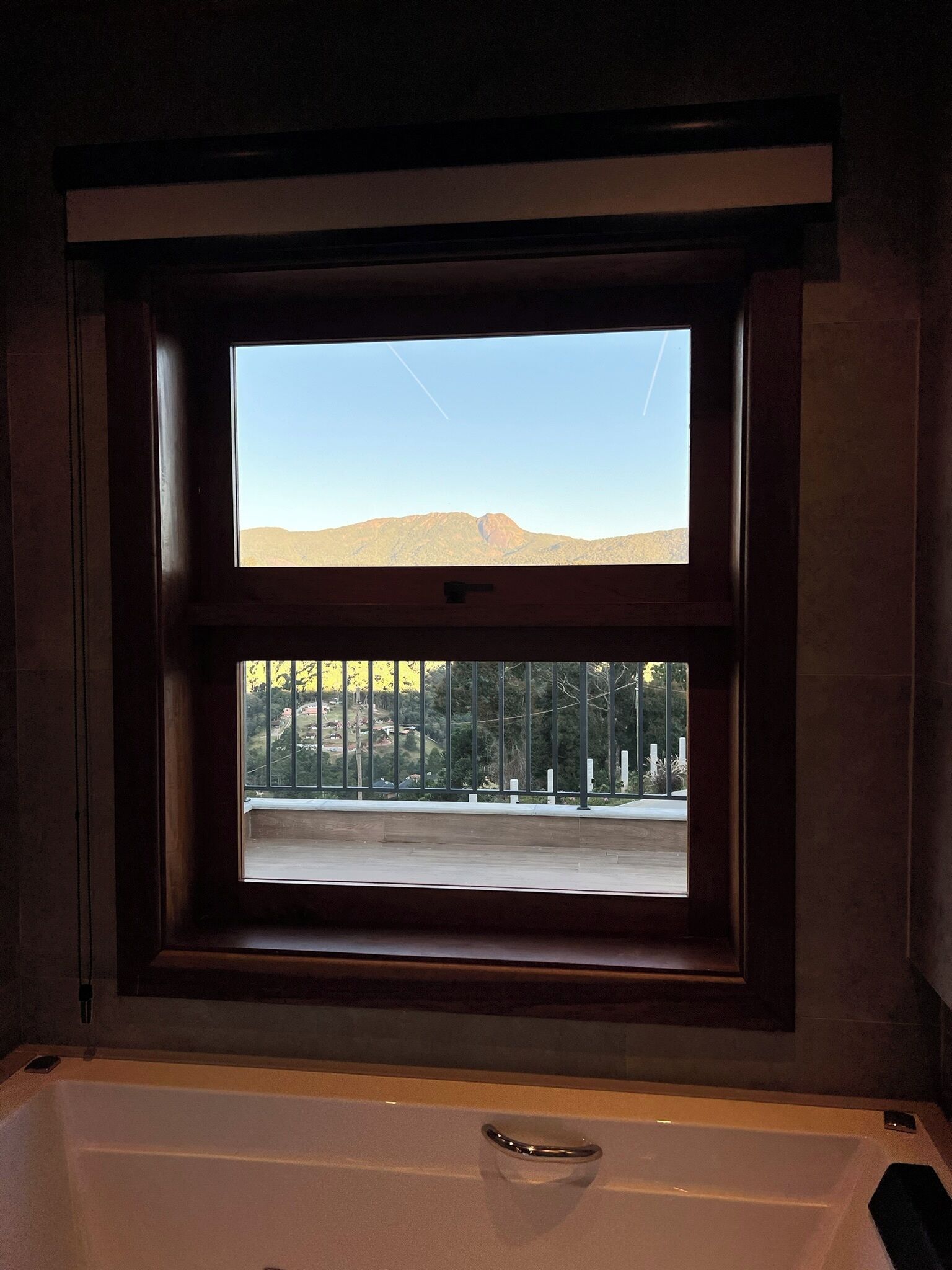 Um banheiro com banheira, janela com vista para uma montanha e vegetação, e guarda-corpo de metal do lado de fora.