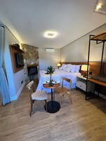 Quarto de hotel com cama, uma pequena mesa com cadeiras, TV montada na parede e uma parede de destaque em pedra.
