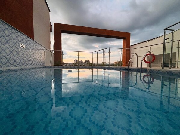 Piscina com piso revestido, rodeada por um corrimão de metal, e vista do horizonte da cidade ao fundo.