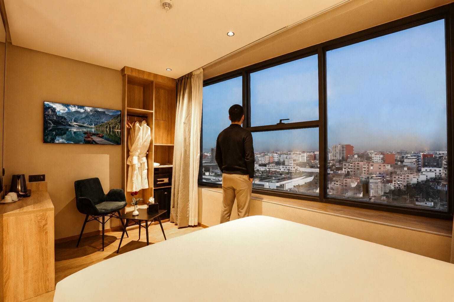 Quarto de hotel com uma janela grande, uma cama, uma televisão, um porta-robes e uma mesa com uma cadeira.