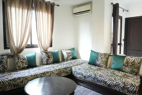 Uma sala de estar com sofá de canto, mesa de centro redonda e janela com cortinas.