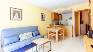 Um espaço de estar compacto com sofá azul, mesa de madeira pequena, área de cozinha com armários de madeira e micro-ondas.