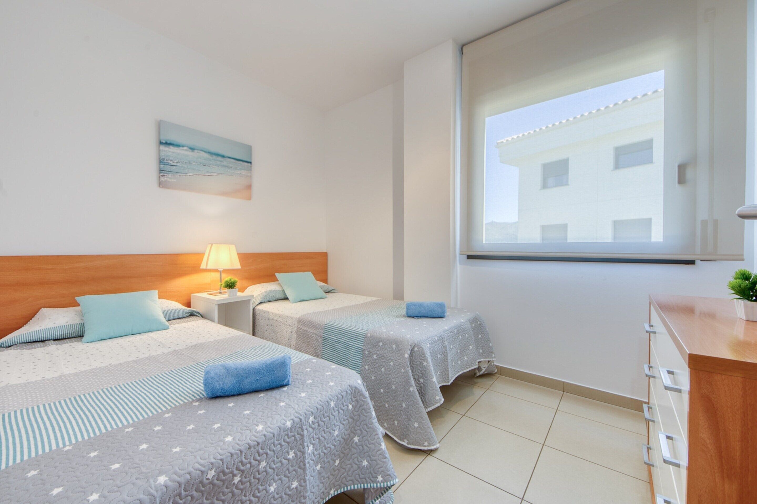 Quarto de hotel com duas camas, uma mesa de cabeceira com abajur, uma janela com vista para um edifício e uma pequena planta no aparador.