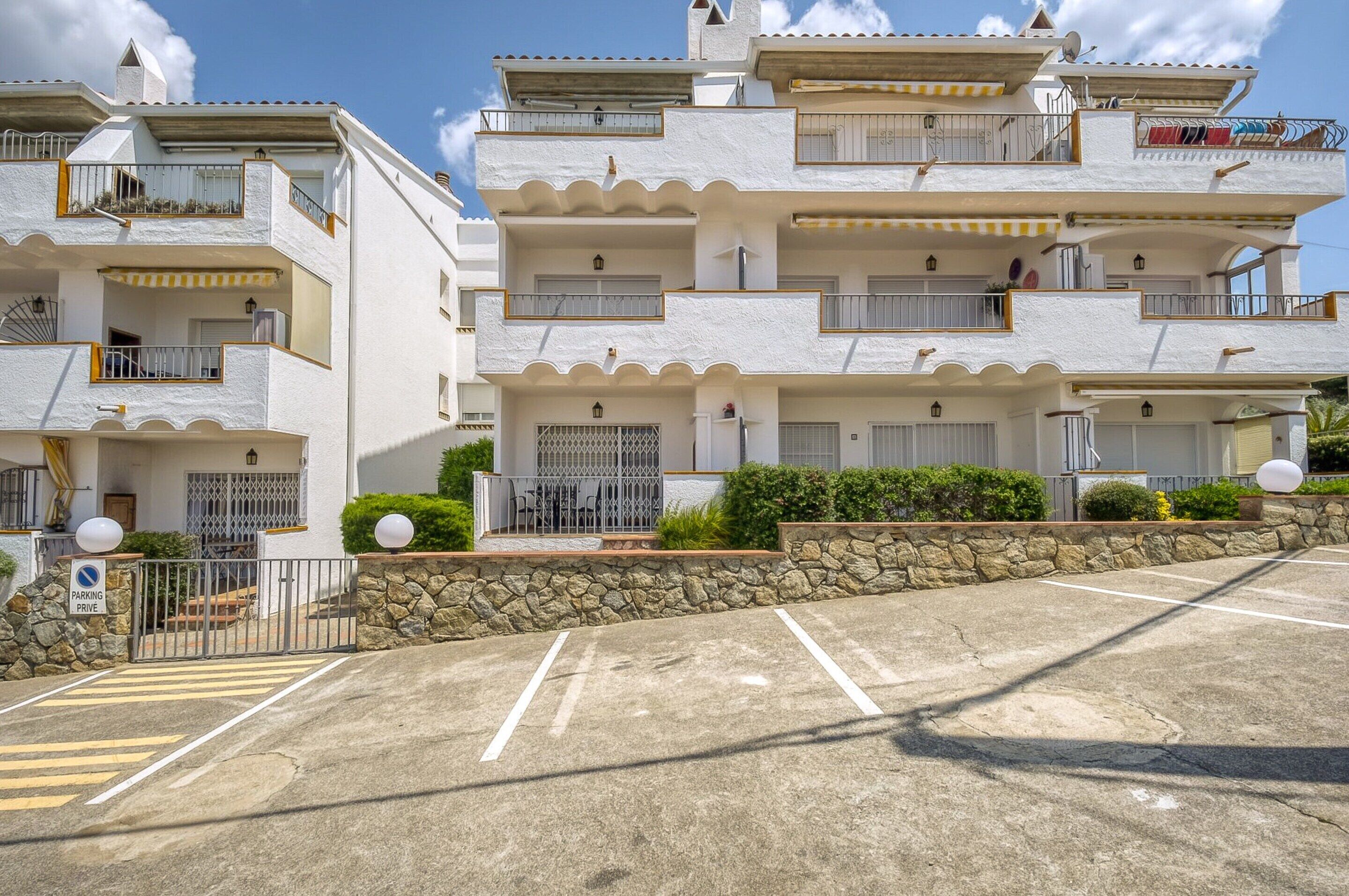 Um edifício residencial com vários andares, varandas, um muro de pedra e uma área de estacionamento.