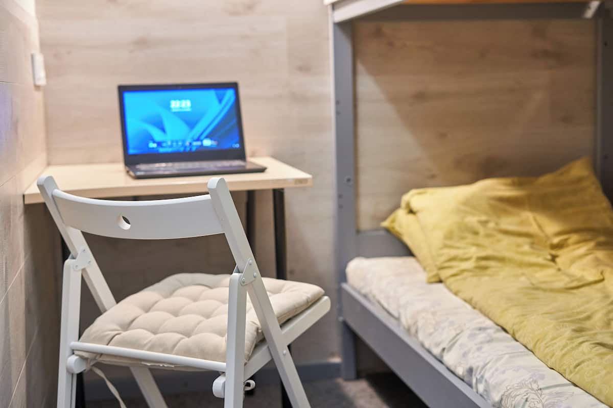 Uma cama de solteiro com um laptop em uma escrivaninha, uma poltrona acolchoada e um travesseiro amarelo.