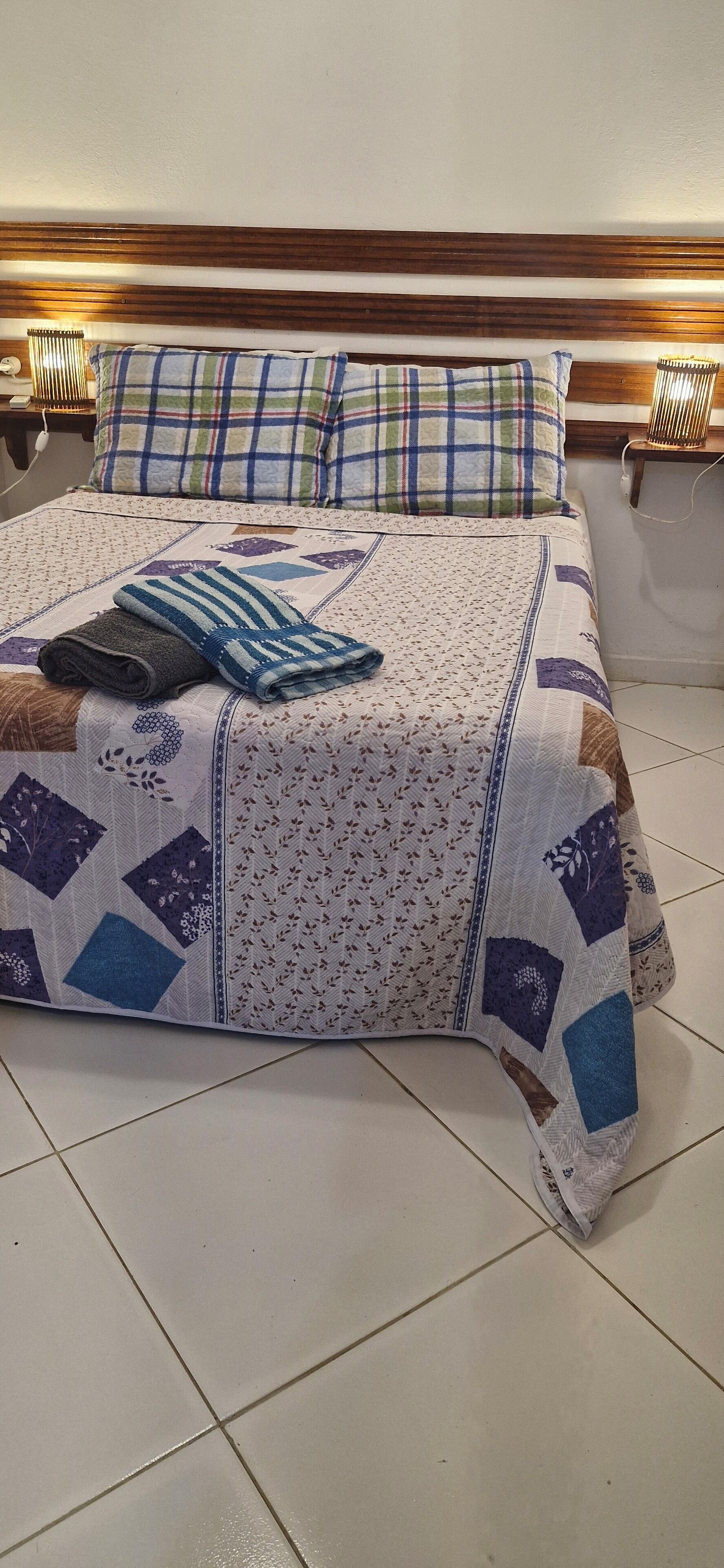 Cama bem arrumada com colcha estampada e almofadas, toalha dobrada e abajur sobre piso de azulejos.