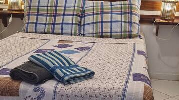 Cama bem arrumada com colcha estampada e almofadas, toalha dobrada e abajur sobre piso de azulejos.