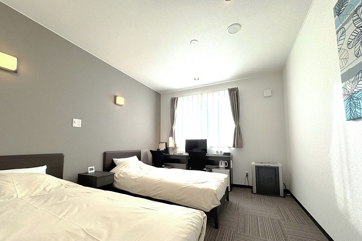 Quarto de hotel com duas camas, uma mesa de trabalho com cadeira, um pequeno aquecedor e uma janela com cortinas.