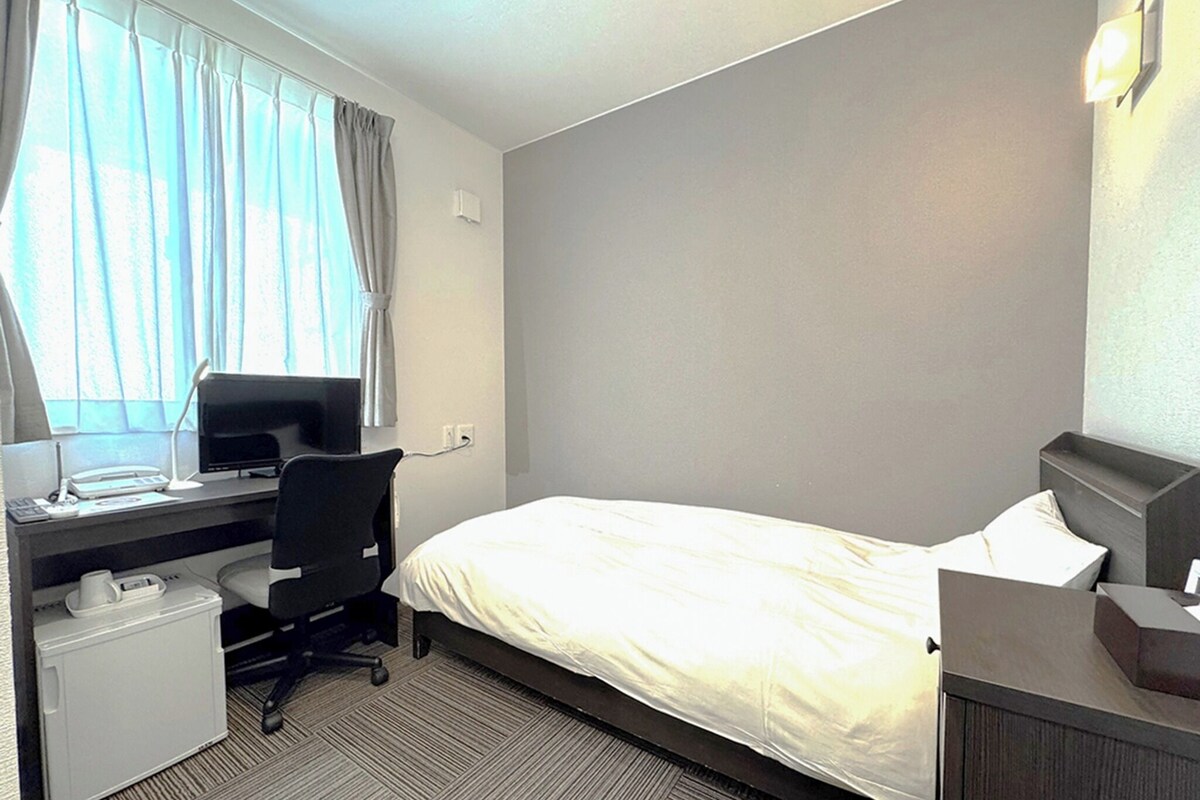 Quarto de hotel com cama, escrivaninha com computador, cadeira, pequeno frigorífico e janela com cortinas.