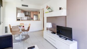 Uma sala de estar moderna com televisão de tela plana, um espaço de jantar com cadeiras de vime e uma cozinha ao fundo.