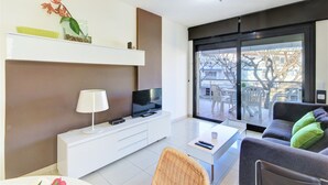 Uma sala de estar moderna com televisão de tela plana, console branco, sofá com almofadas verdes e uma grande janela com vista para o exterior.