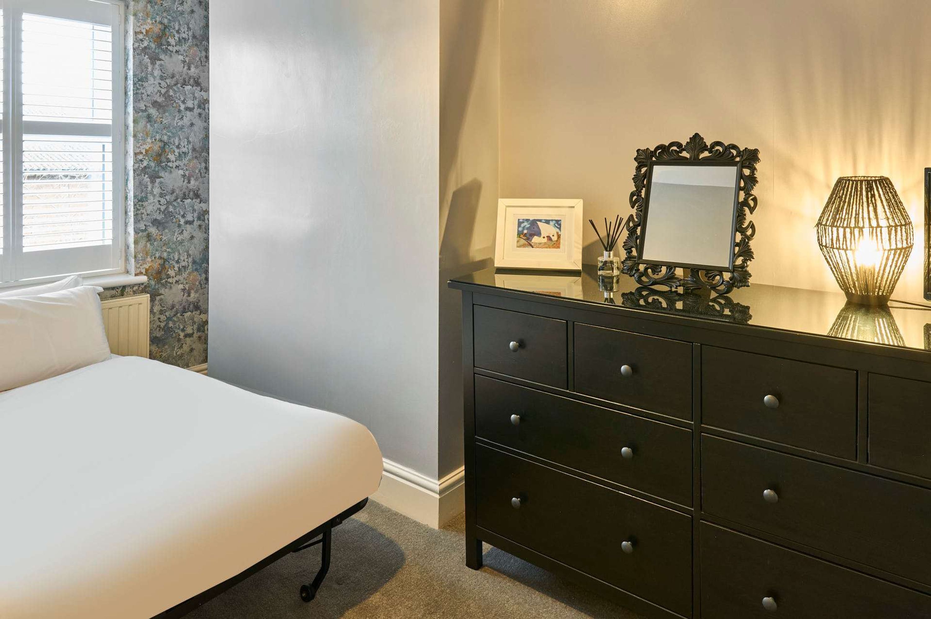 Quarto com cama, cómoda com gavetas, espelho, moldura de quadro e abajur.