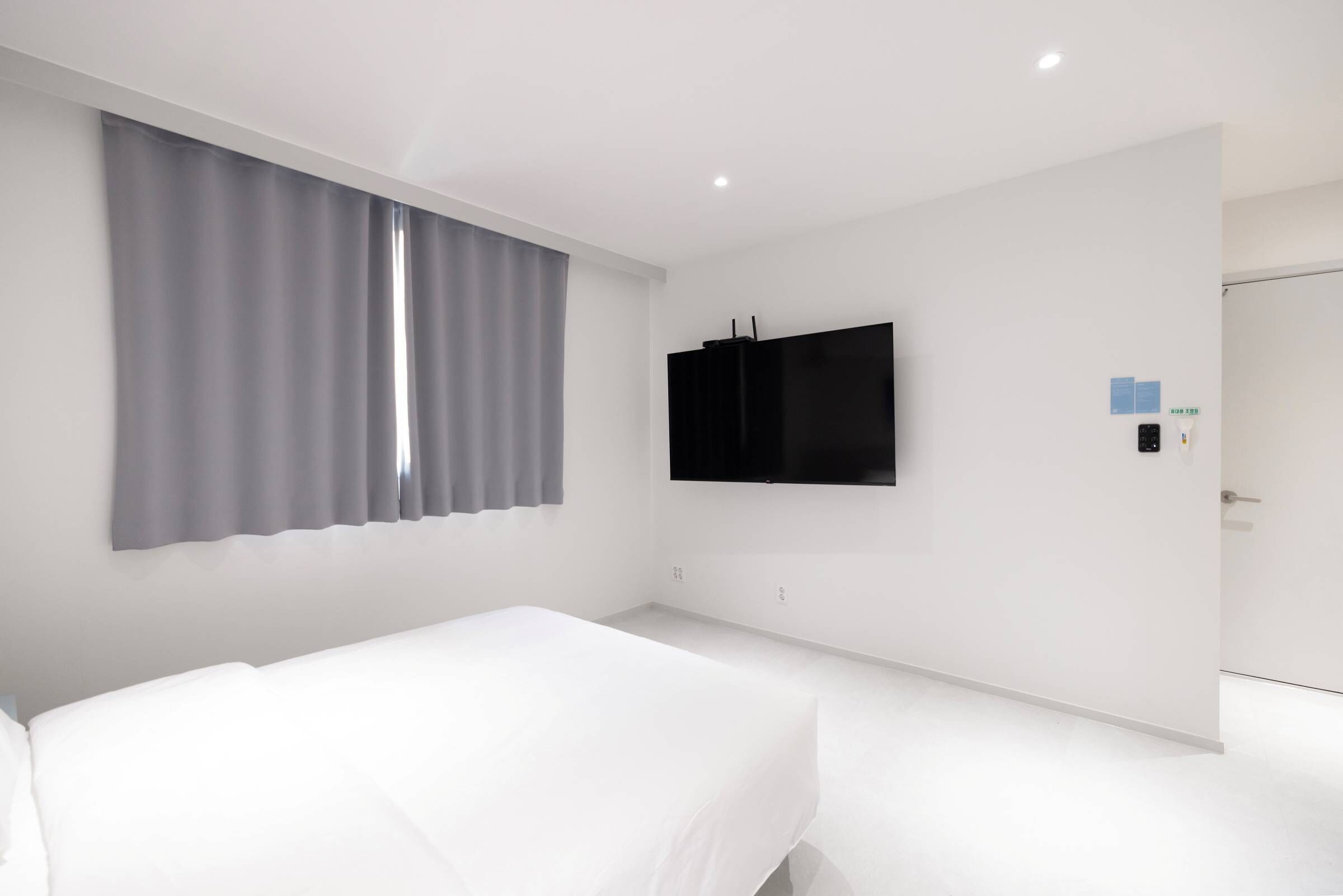 Quarto de hotel moderno com uma cama grande, uma TV de tela plana na parede e uma janela com cortinas cinzentas.
