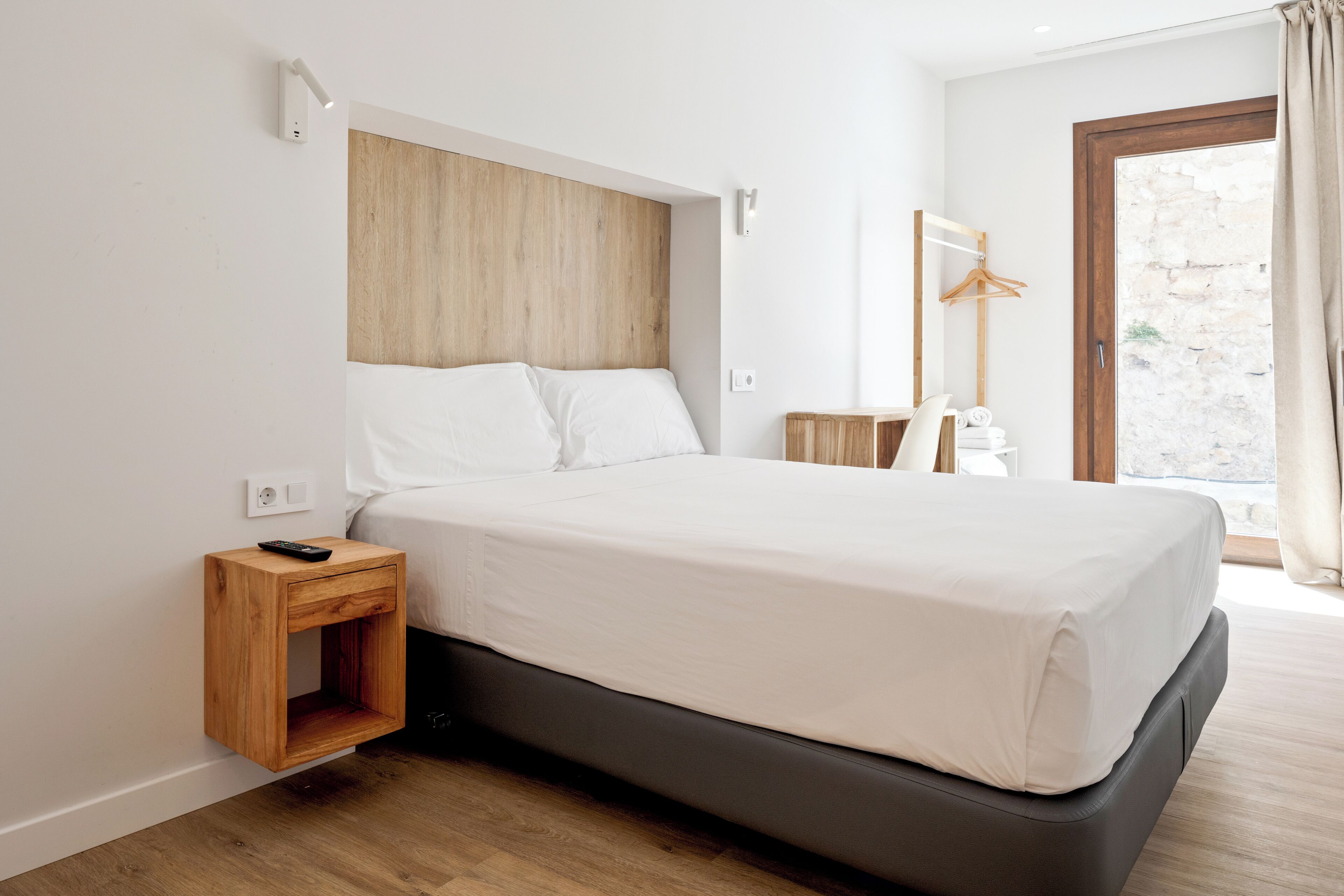 Quarto de hotel moderno com cabeceira de madeira, uma cama de solteiro, uma pequena mesa de cabeceira de madeira e uma ampla janela com cortinas.